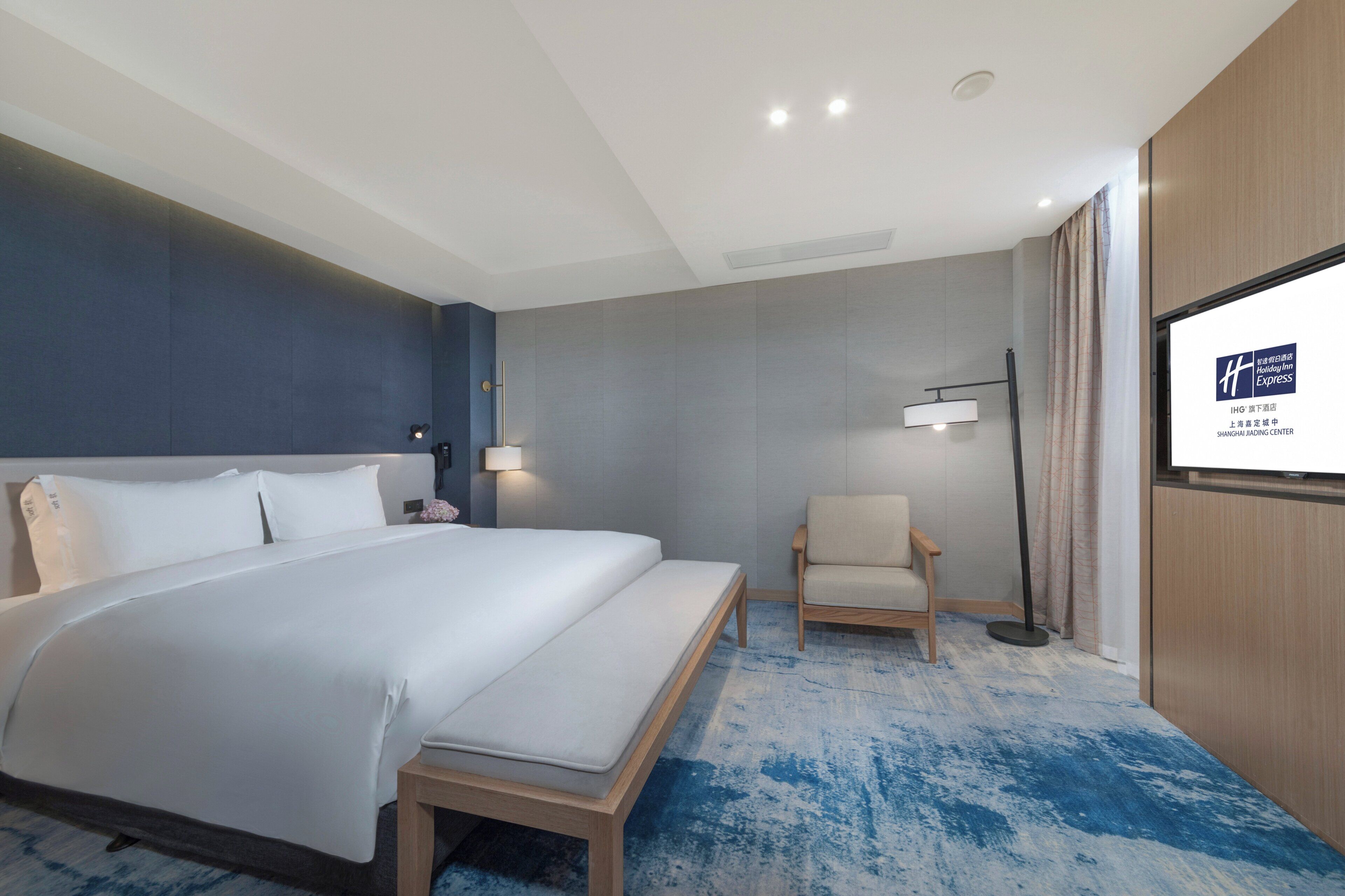 Quarto de hotel com uma cama grande, mesa de cabeceira, uma cadeira e uma TV exibindo o nome do hotel.