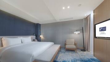 Quarto de hotel com uma cama grande, mesa de cabeceira, uma cadeira e uma TV exibindo o nome do hotel.