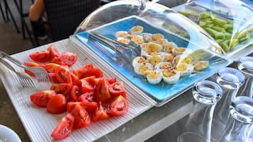 Uma mesa de buffet com fatias de tomate e ovos cozidos em um prato branco, e um recipiente de vidro com vegetais verdes.