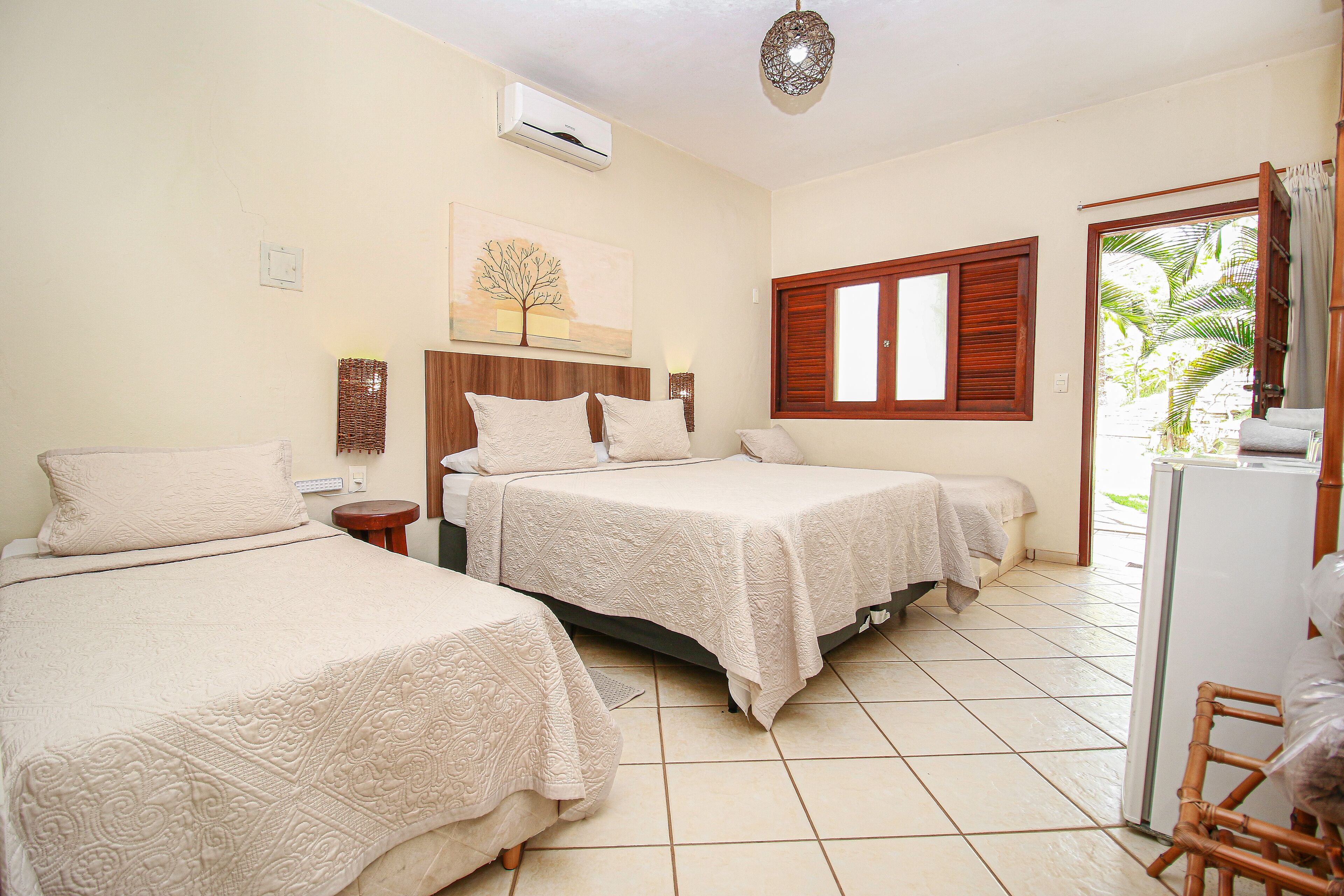 Quarto de hotel com duas camas, cabeceira de madeira, ar condicionado fixado na parede, luminária no teto e janela com persianas de madeira.