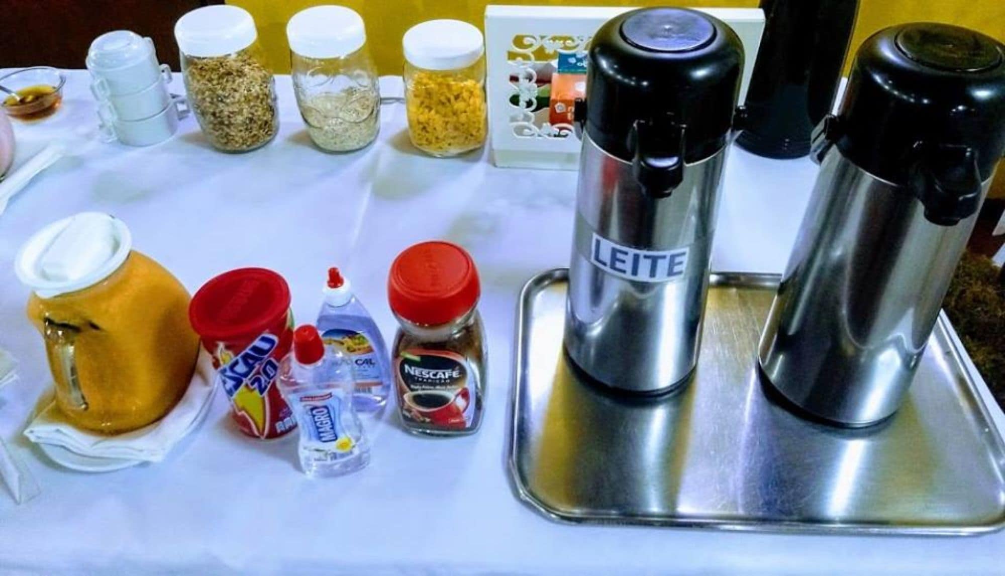 Uma mesa com diversos alimentos e acompanhamentos, incluindo potes de temperos, uma garrafa de Nescafé e duas garrafas térmicas de aço inoxidável com a etiqueta 