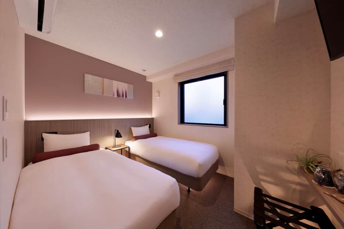 Quarto de hotel com duas camas, uma janela, uma obra de arte na parede e uma pequena mesa com abajur.