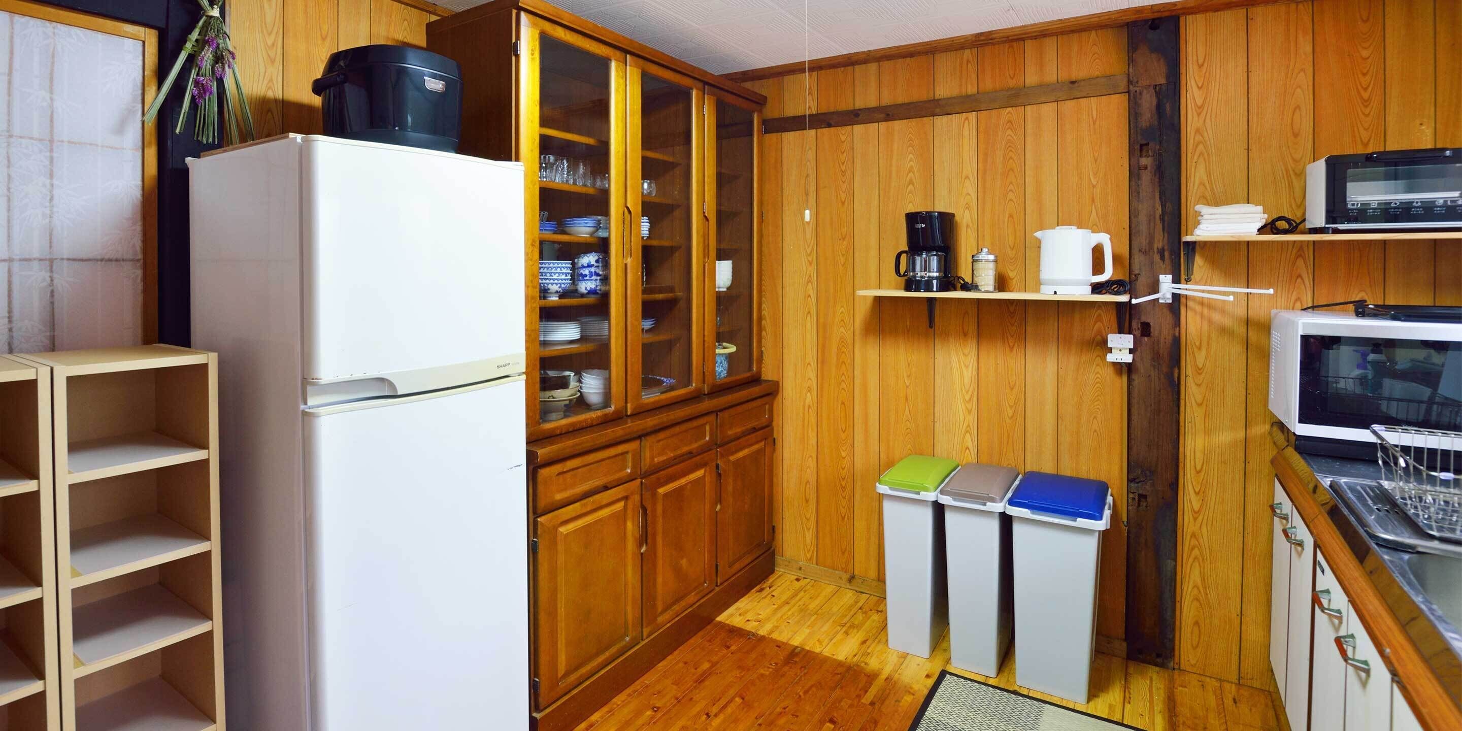 Uma cozinha compacta com armários de madeira, uma geladeira branca, micro-ondas e cafeteira em uma prateleira. Há três lixeiras e uma pequena estante com louças.