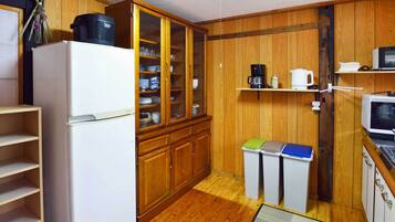 Uma cozinha compacta com armários de madeira, uma geladeira branca, micro-ondas e cafeteira em uma prateleira. Há três lixeiras e uma pequena estante com louças.