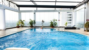 Piscina interior com vista clara da cidade, rodeada por plantas em vasos e uma espreguiçadeira.