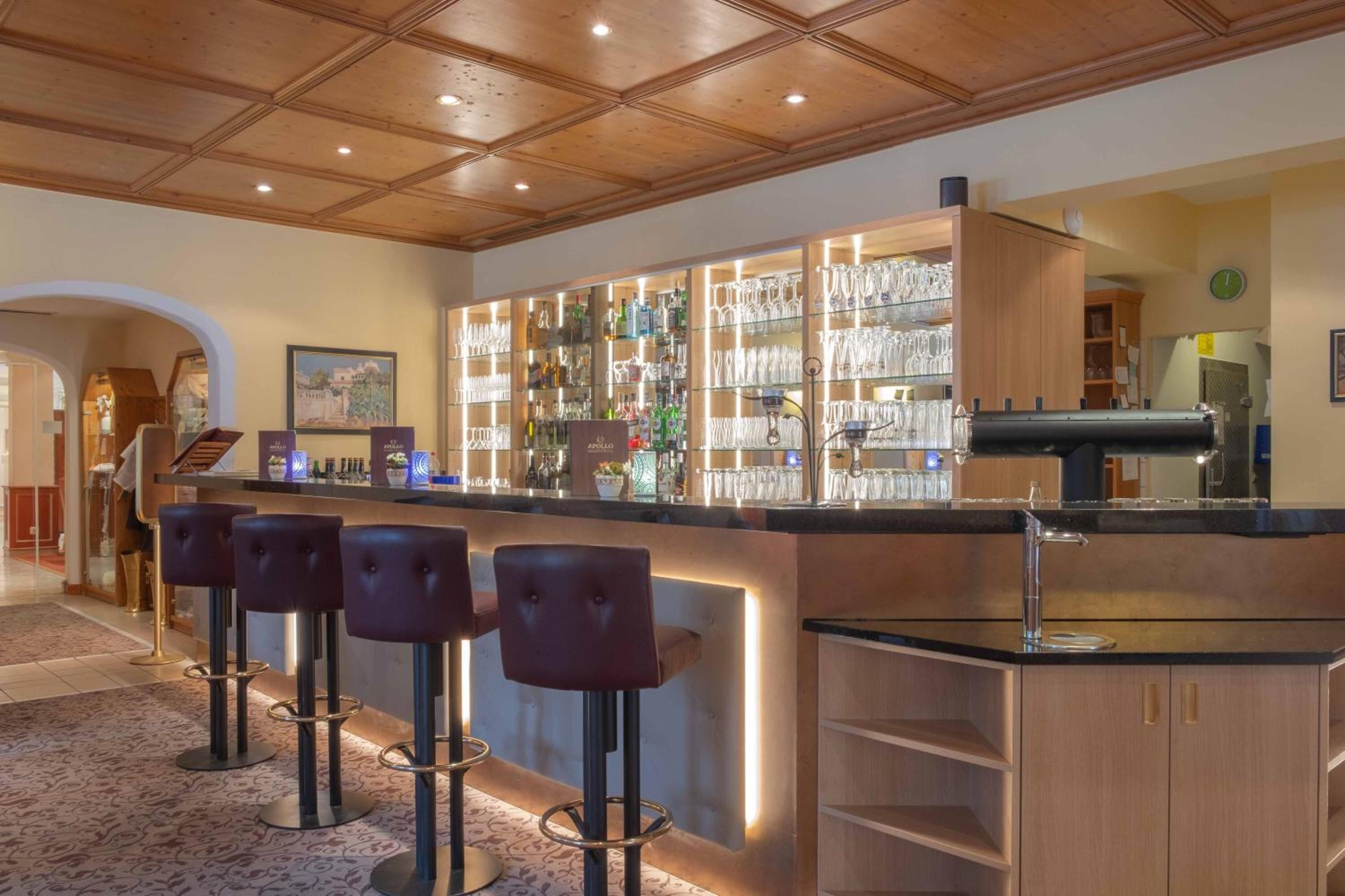 Uma área de bar com teto de madeira, banquetas e estantes cheias de garrafas e copos.