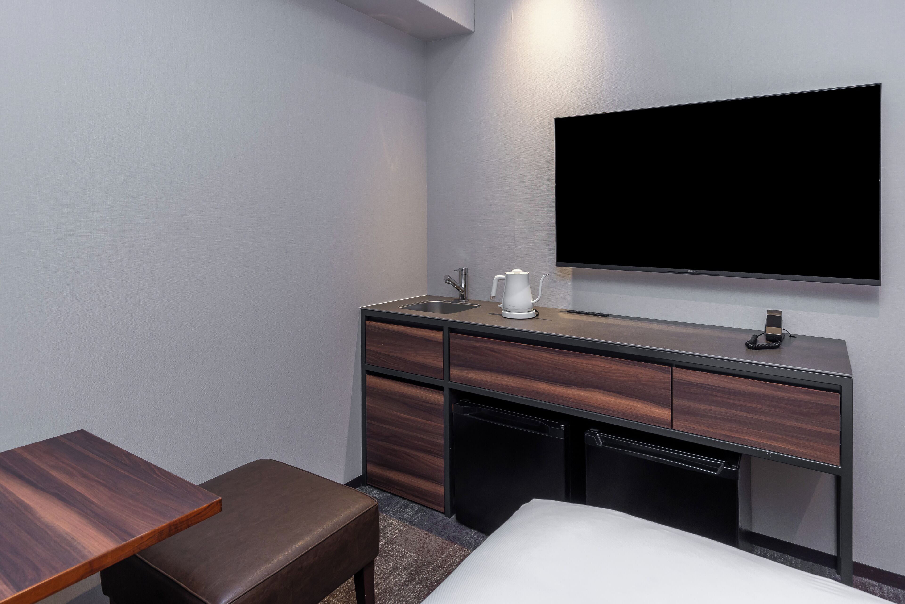 Quarto de hotel moderno com escrivaninha de madeira, um banco, lavatório embutido e televisão de tela plana fixada na parede.