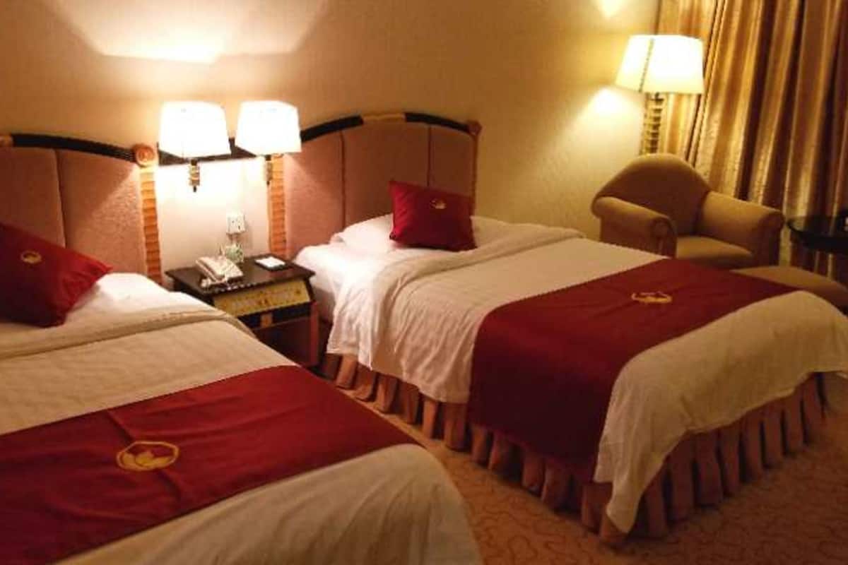 Quarto de hotel com duas camas, cada uma com colcha vermelha e almofada combinando. Há duas luminárias de cabeceira, uma mesa de cabeceira com telefone, e uma cadeira no canto.
