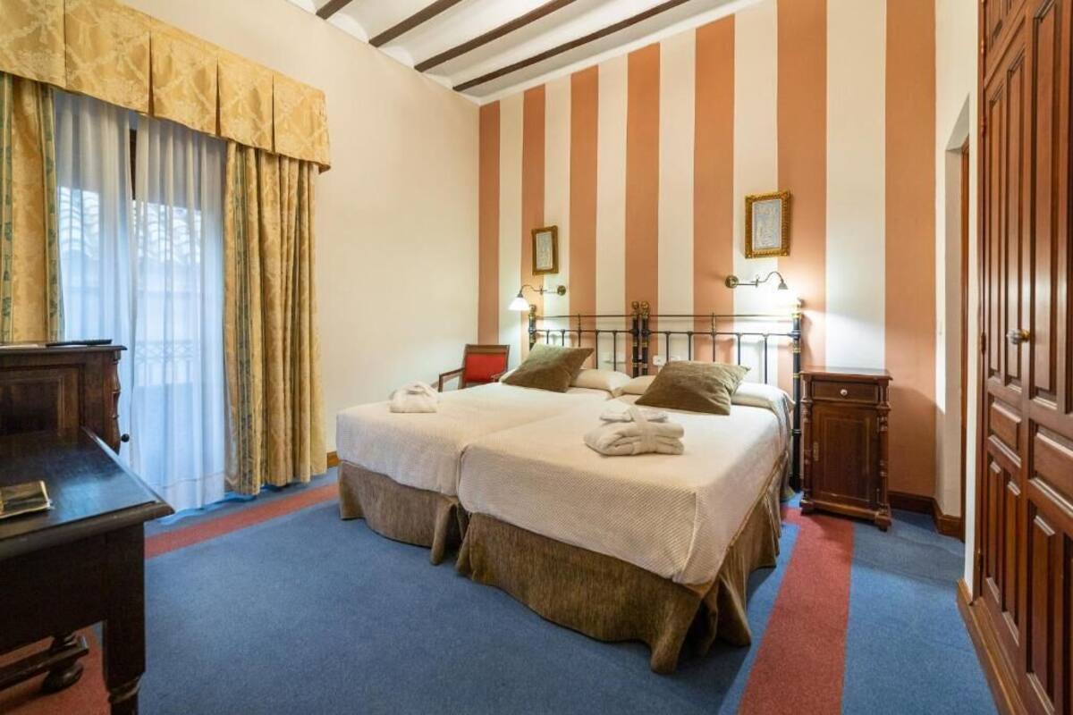 Quarto de hotel com paredes listradas, duas camas, uma escrivaninha e um guarda-roupa.