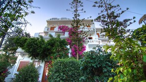 Um hotel com fachada branca, varandas e plantas floridas em tons de rosa.