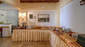 Disposição de buffet com uma mesa comprida coberta por uma toalha bege, diversos alimentos e um espaço para café.