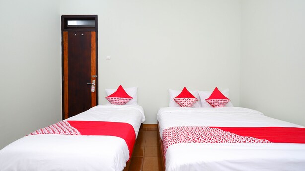 Quarto com duas camas, cada uma com roupa de cama branca e detalhes vermelhos, uma porta de madeira e uma parede branca.
