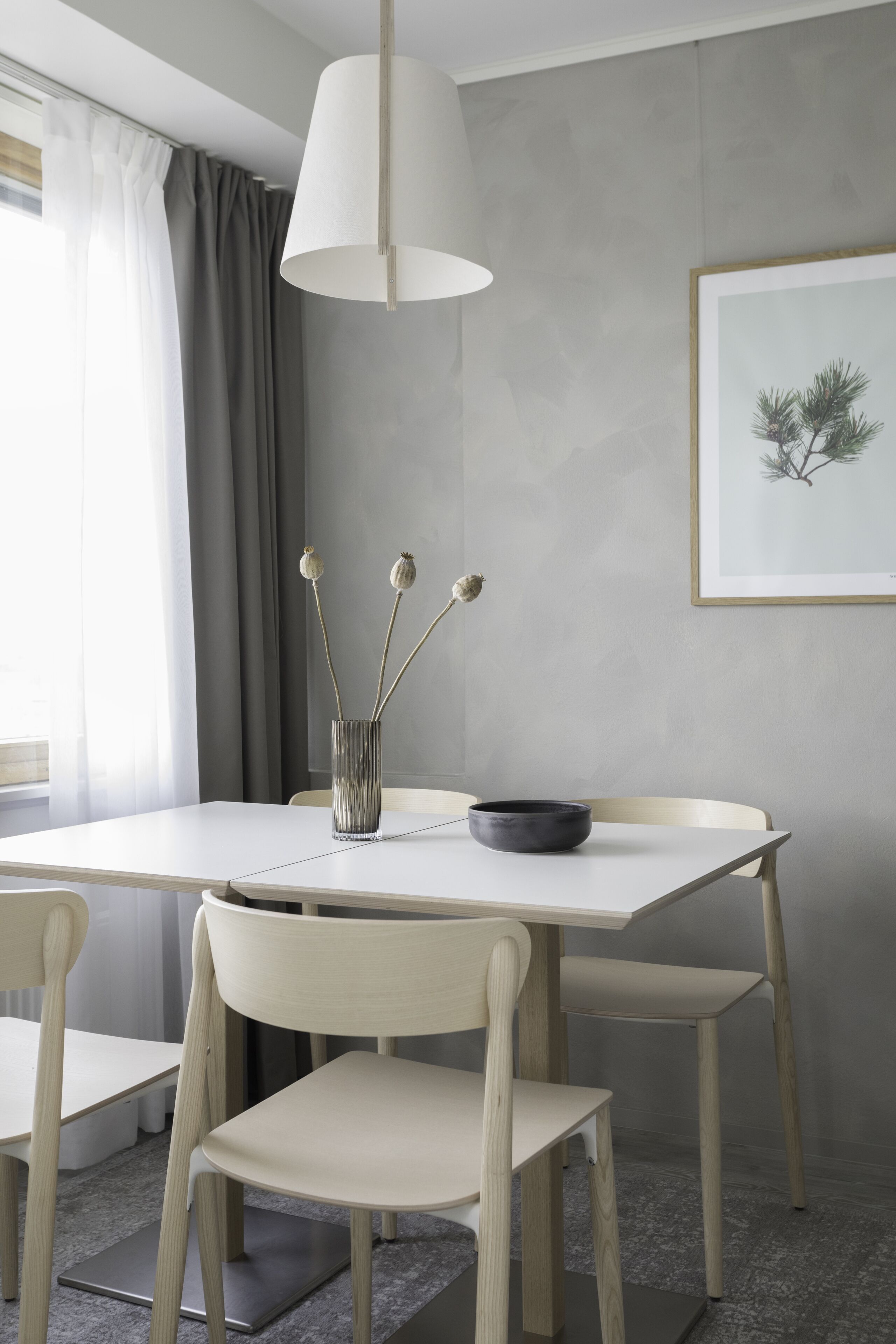 Uma zona de refeições moderna com uma mesa branca, duas cadeiras, um vaso com flores secas e uma ilustração botânica emoldurada na parede.