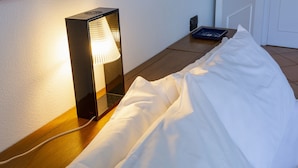 Uma mesa de cabeceira com uma luminária, um controle remoto e um travesseiro branco sobre um piso de madeira.