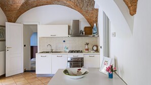 Cozinha com armários brancos, painel de azulejos, arco de tijolos e um espaço de refeições com mesa e um recipiente decorativo.