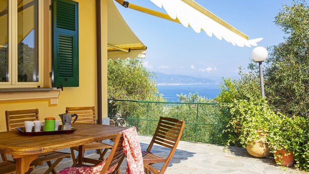 Um pátio com mesa e cadeiras, vista para o mar e um edifício amarelo com persianas verdes.