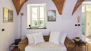 Quarto com um sofá branco, uma cadeira de vime, uma mesa pequena e uma janela com vista para o verde.