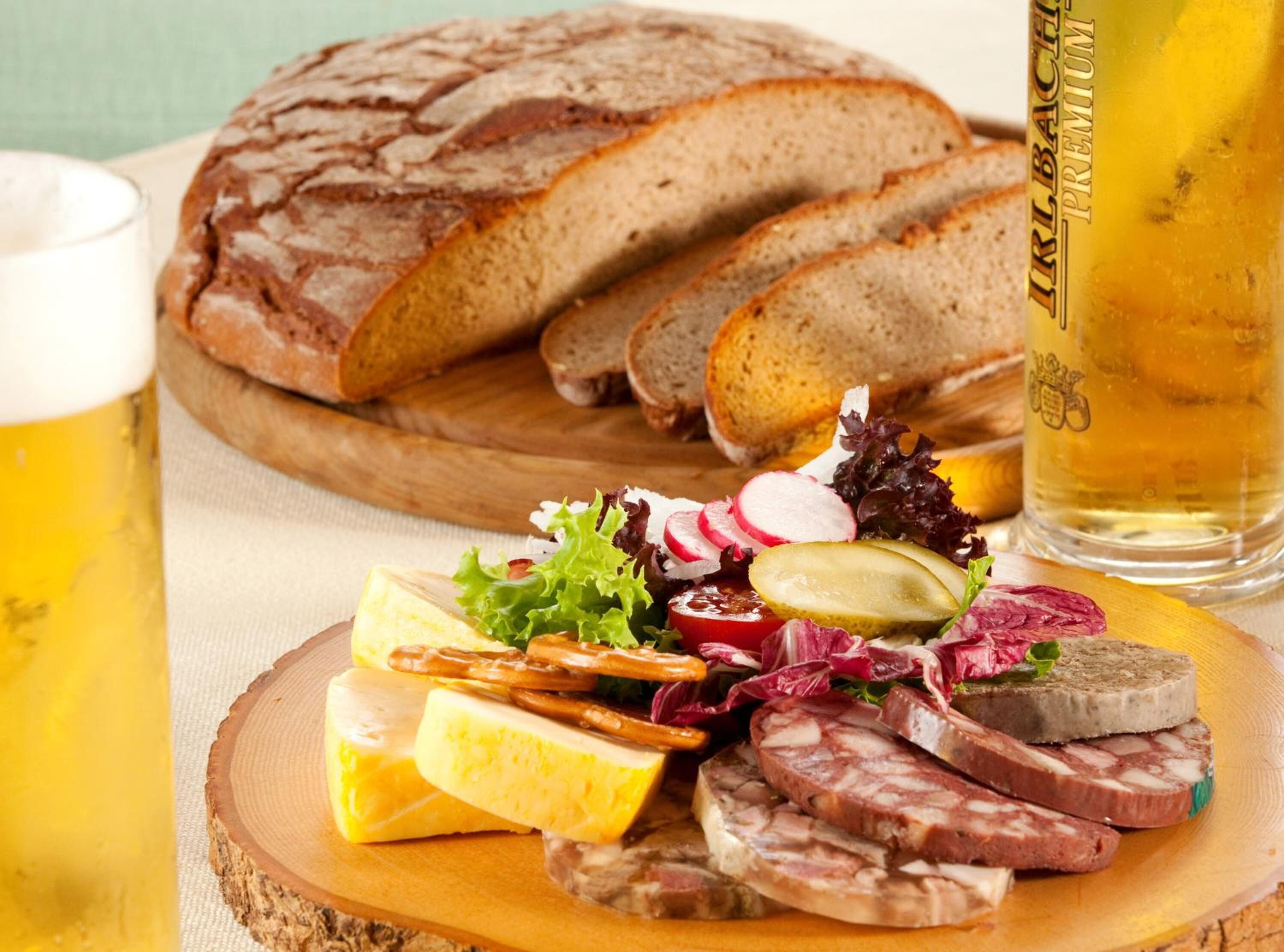 Tábua de madeira com fatias de carne, queijo e legumes, acompanhada de fatias de pão e um copo de cerveja.