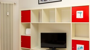 Quarto com televisão sobre suporte, radiador e estantes em vermelho e branco.