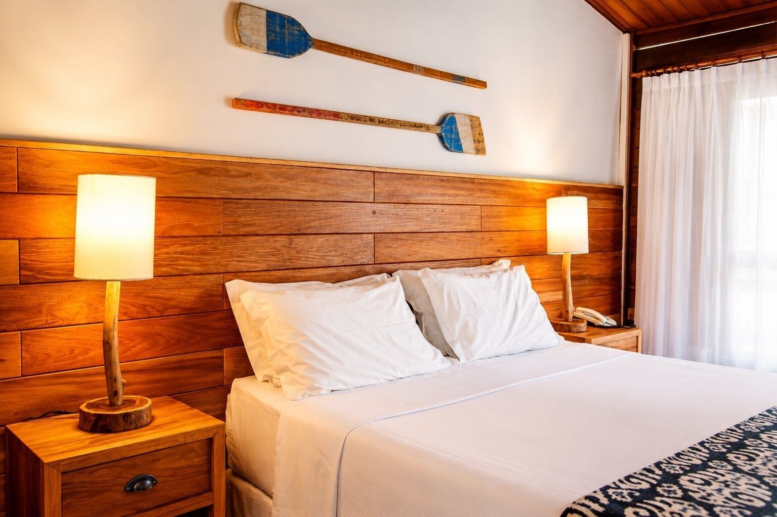 Quarto de hotel com cabeceira de madeira, duas luminárias de apoio, cama com roupa de cama branca e manta estampada.