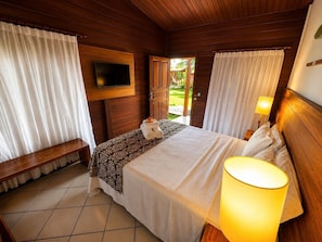 Quarto de madeira com uma cama, um banco, uma TV e uma luminária.