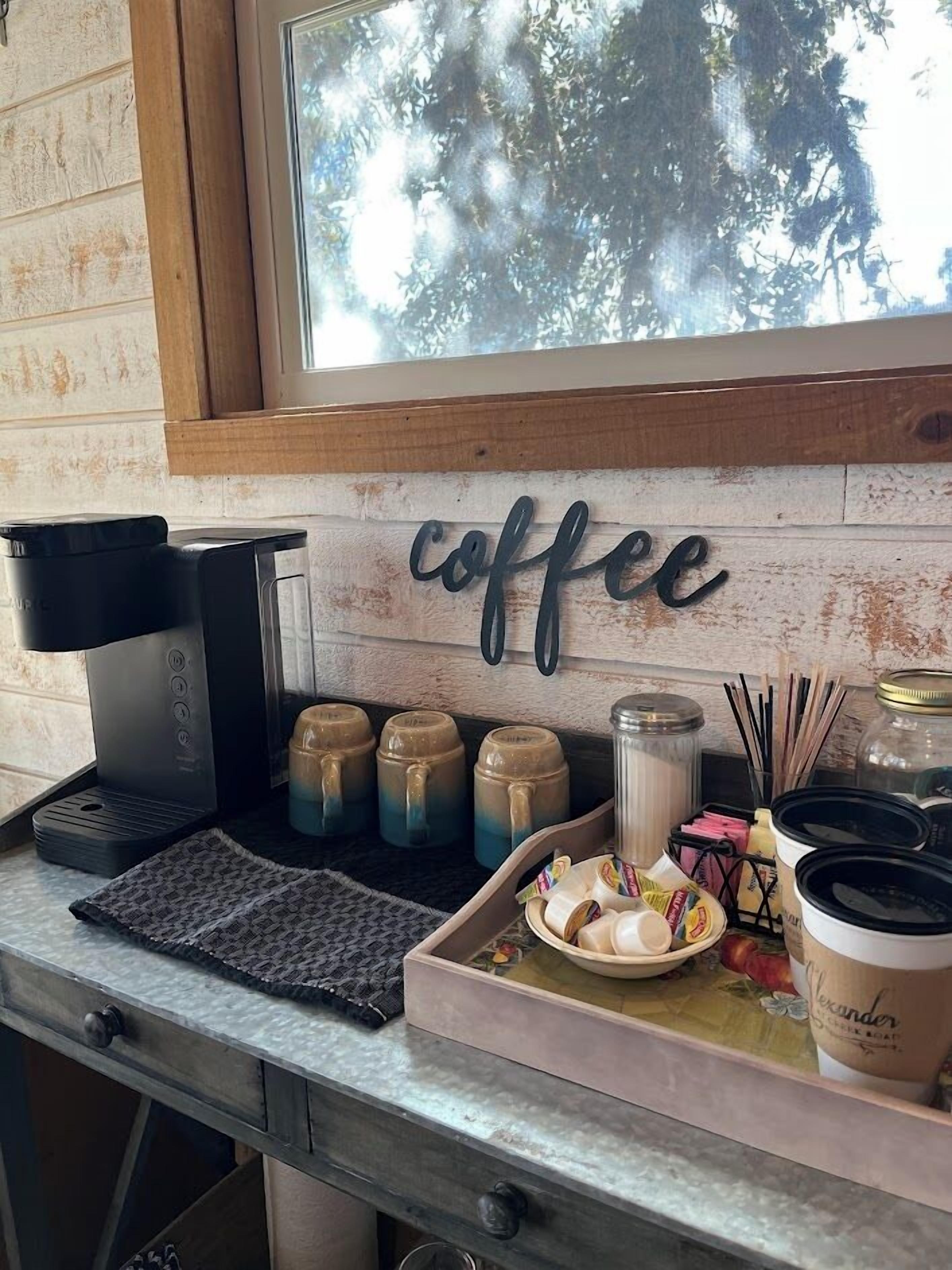 Estação de café com cafeteira, canecas e bandeja com xícaras e acompanhamentos.