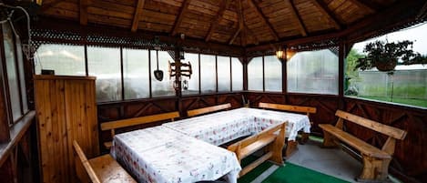 Uma área de estar ao ar livre coberta, com bancos e mesas de madeira, um tapete verde e teto de madeira.