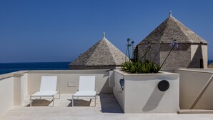 Terraço no telhado com duas poltronas brancas, uma pequena planta em vaso e um edifício com telhado pontiagudo.