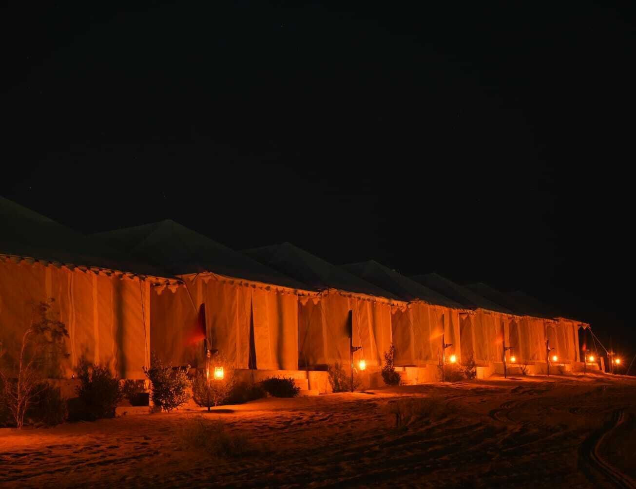 Um edifício amplo e iluminado, com várias janelas e telhado plano, rodeado por vegetação rala.