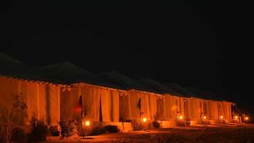 Um edifício amplo e iluminado, com várias janelas e telhado plano, rodeado por vegetação rala.