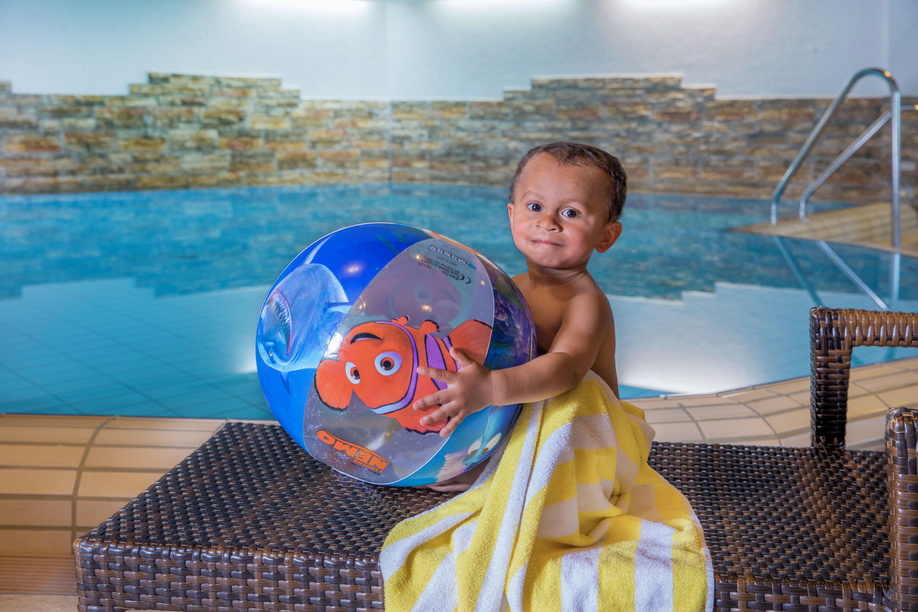 Criança sentada em uma cadeira de vime perto de uma piscina interna, com um muro de pedra ao fundo.
