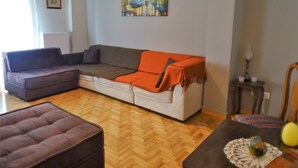 Uma sala de estar com um sofá seccional, um pequeno banquinho, uma mesa de centro e um quadro na parede.