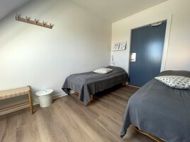 Quarto com duas camas, um banco de madeira, uma lixeira branca e uma porta azul.