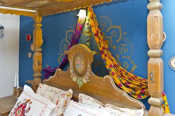 Uma cama de madeira com peseira entalhada, parede azul com desenhos decorativos e tecido amarelo e vermelho pendurado sobre a peseira.