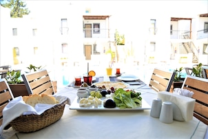 Uma mesa posta com diversos alimentos, incluindo pão, queijo, azeitonas e verduras, com vista para edifícios ao fundo.