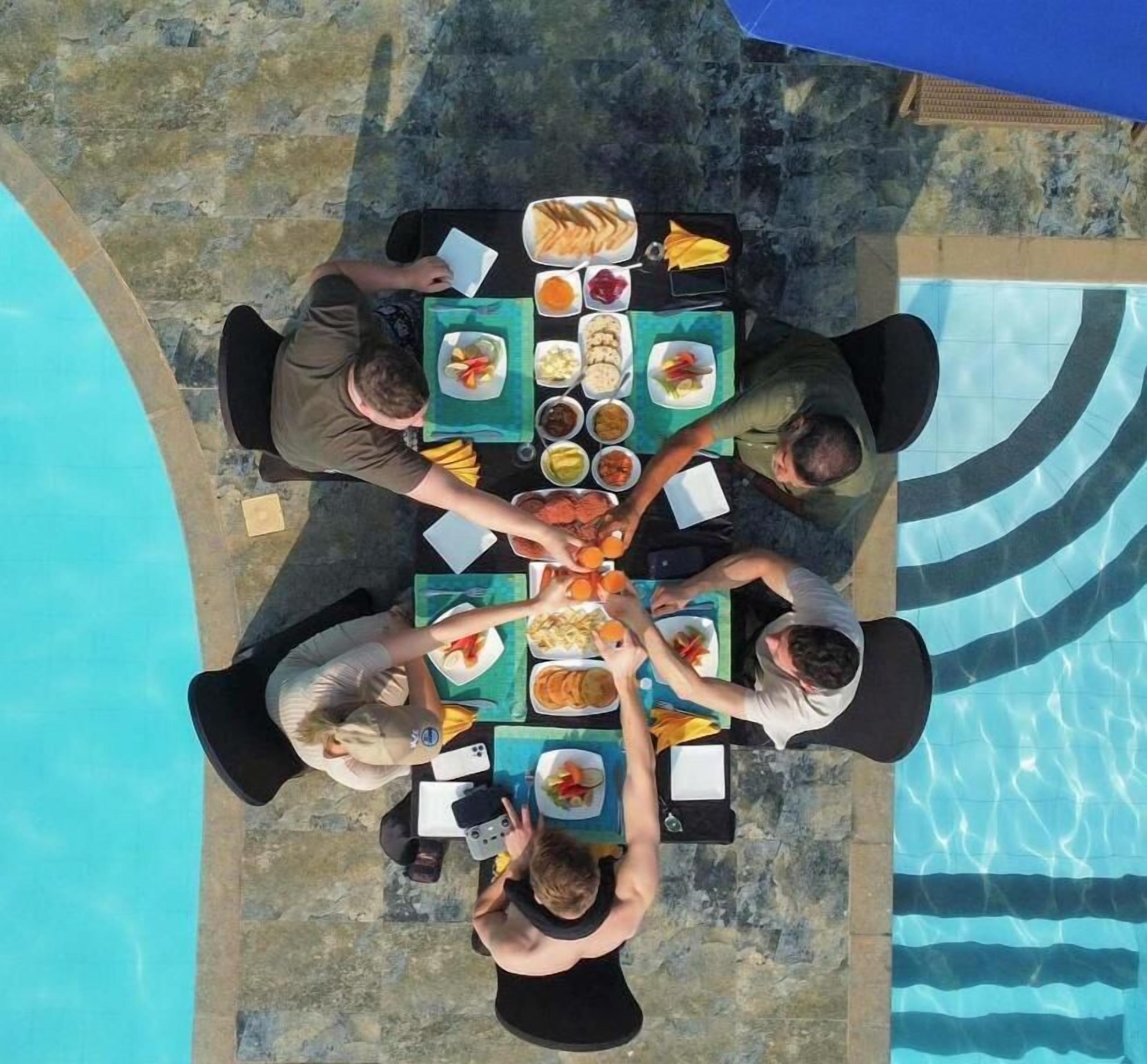 Um grupo de pessoas perto de uma mesa com comida, próximo a uma piscina.