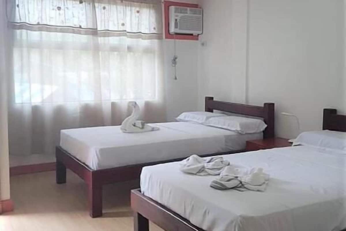 Quarto de hotel com duas camas de solteiro, uma janela com cortinas finas, uma luminária no teto e um ar-condicionado fixado na parede.