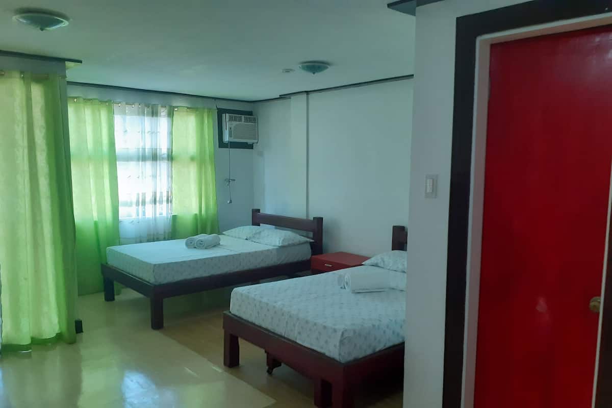 Quarto com duas camas, uma porta vermelha e uma janela com cortinas verdes.