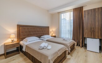 Quarto de hotel com duas camas, cabeceira de madeira, uma pequena escrivaninha com luminária, mini-geladeira e uma janela grande com cortinas.