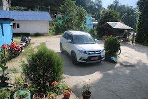 Um SUV branco estacionado em área rural, com uma trilha de terra, cercado por vasos de plantas e um pequeno edifício azul.