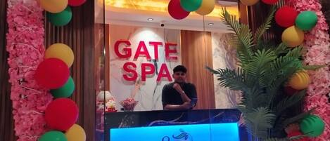 Entrada de um spa, com uma mesa de recepção, uma placa com o texto "GATE SPA" e um arco decorado com balões.
