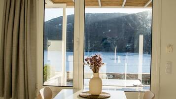 Quarto com uma mesa e duas cadeiras, janela com vista para um corpo d'água e montanhas, e um aparelho de ar condicionado.