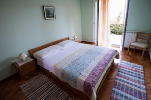 Quarto com cama de madeira, mesa de cabeceira com luminária, uma cadeira e vista para o exterior por uma porta deslizante.