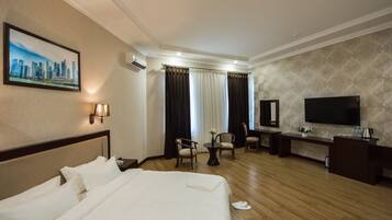 Quarto de hotel com uma cama grande, uma escrivaninha com televisão de tela plana, um espaço de estar com cadeiras e uma pintura da paisagem urbana na parede.