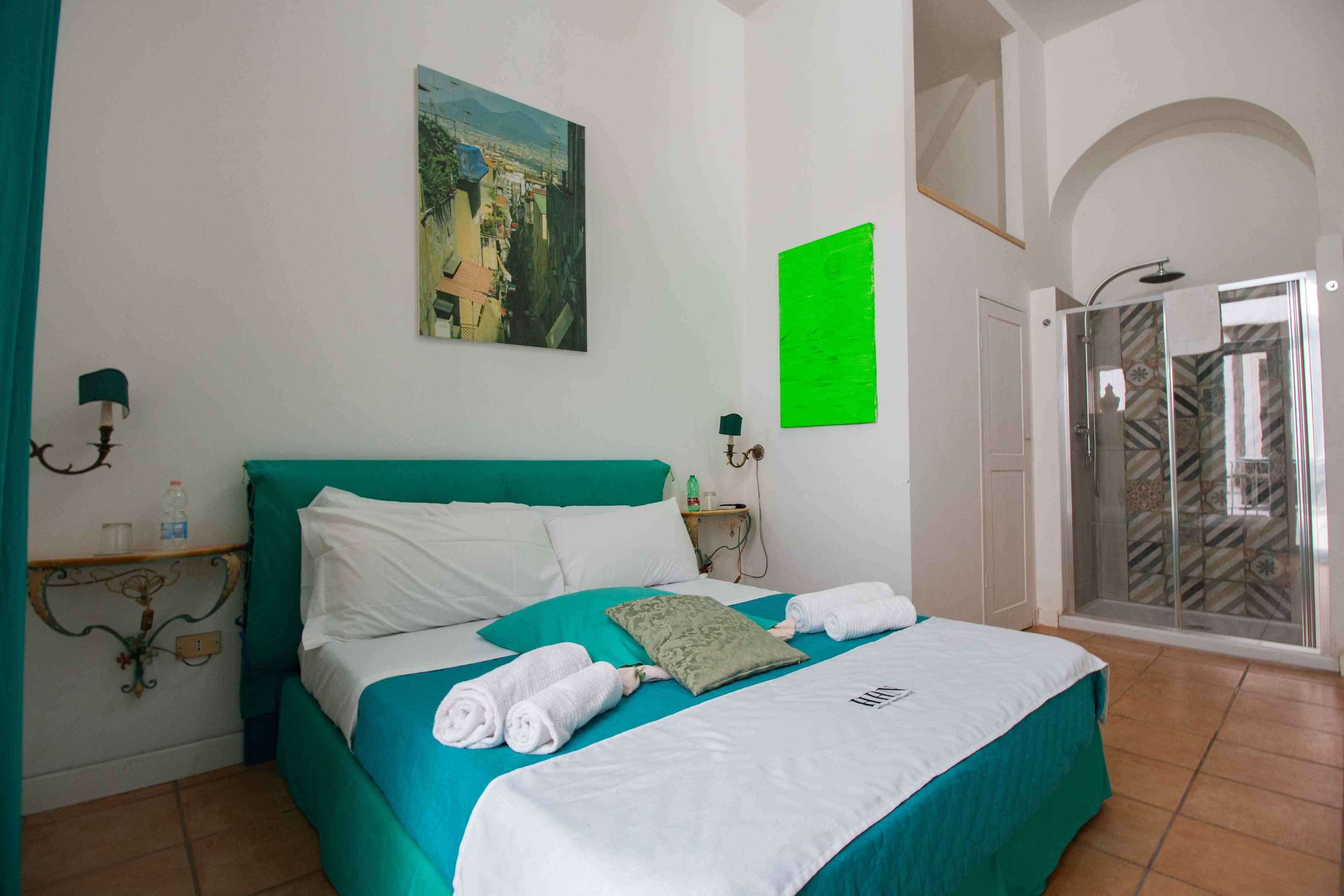 Quarto com cama, parede verde, quadro, placa verde, chuveiro e piso ladrilhado.