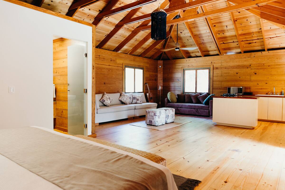 Interior acolhedor de uma cabana de madeira, com uma cama, dois sofás, uma cozinha e uma mesa de jantar.