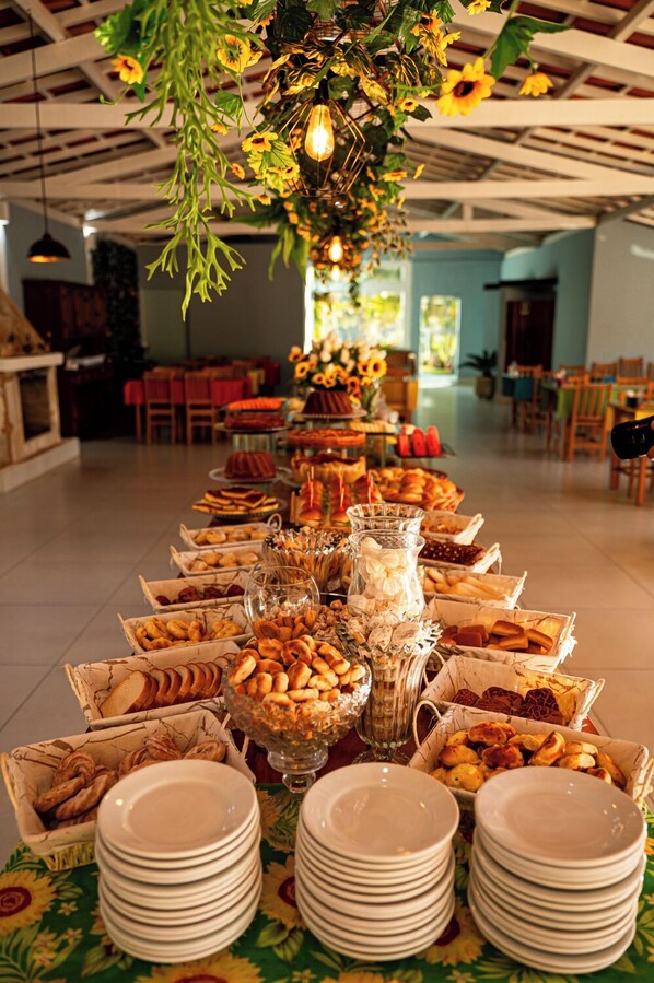 Mesa de buffet com diversos alimentos e pratos, num espaço com pé-direito alto e plantas penduradas.