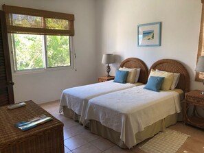 Quarto com duas camas, uma mesinha de vime, janela com persianas e um quadro na parede.