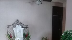 Quarto com ventoinha no teto, um ornamento branco na parede, sofá roxo e plantas em vasos.