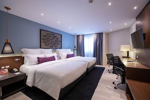 Quarto de hotel com duas camas, uma escrivaninha, uma cadeira, uma televisão e um quadro na parede.
